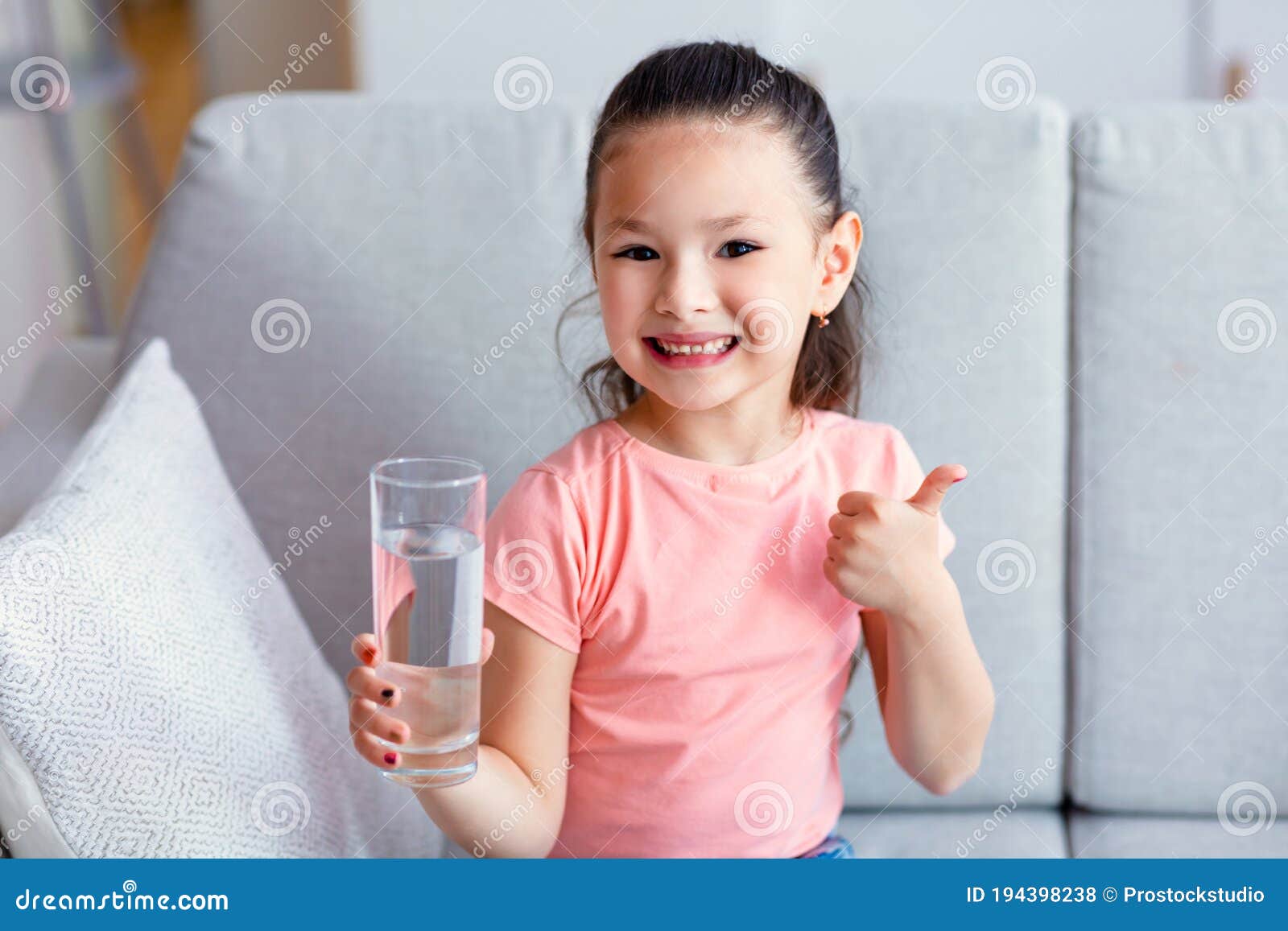 Chinese Girl Holding Glass Of Water Gesturing Thumbs Up Sitting Indoors