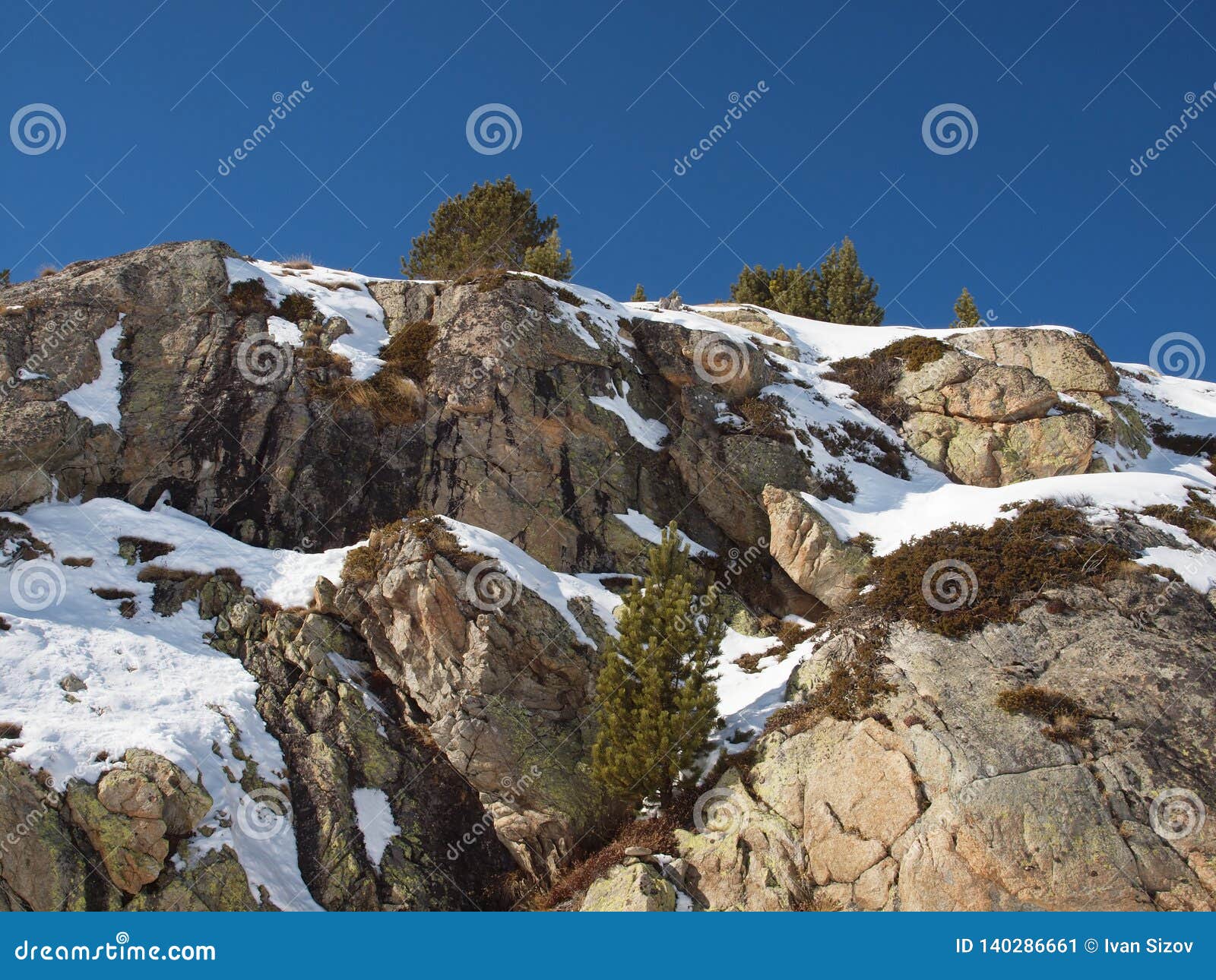 A Cliff on a Snowy Slope Against the Blue Sky with a Lone Tree on Top ...