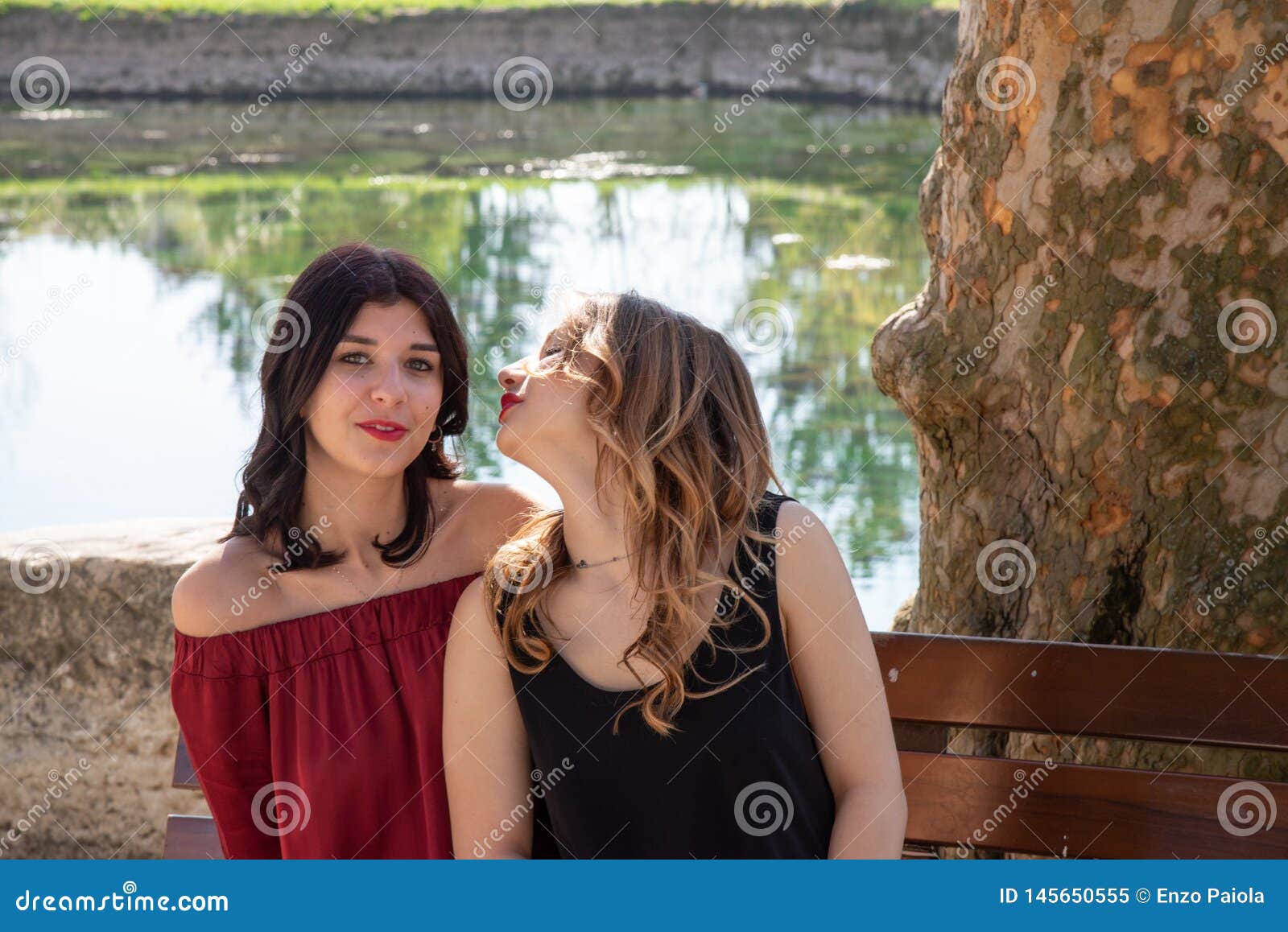 Close Up Of Two Friends Sitting On A Bench By A Lake A Blonde The