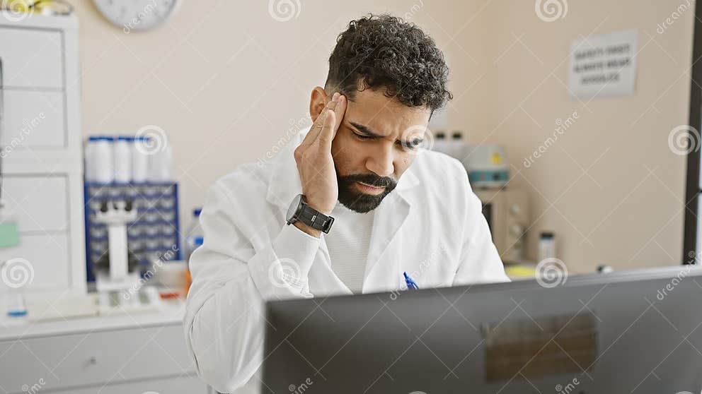 A Concerned Young Man in a White Lab Coat Studies a Computer Screen in ...