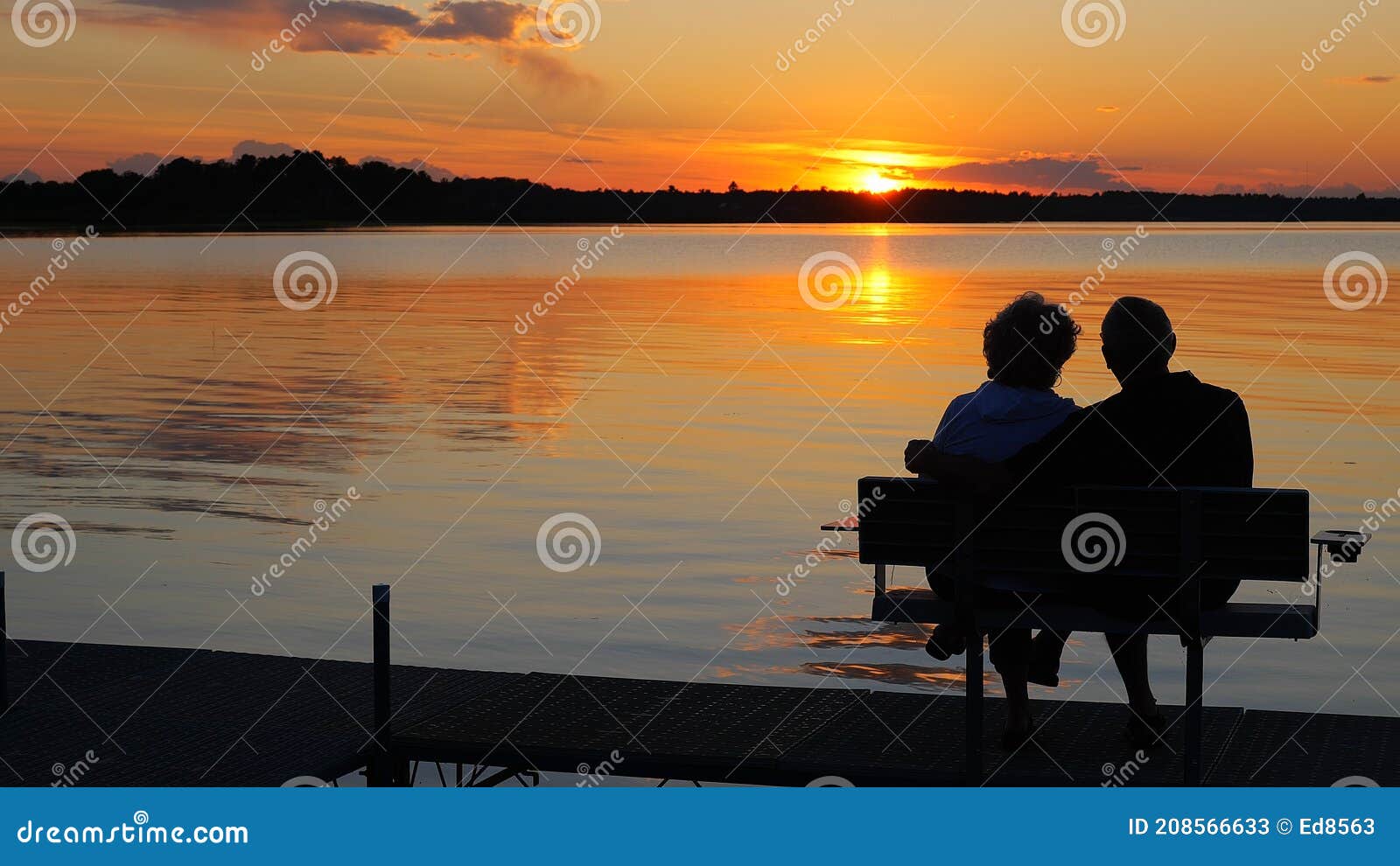 A Couple On A Bench On A Dock Enjoy A Sunset Over A Beautiful Lake