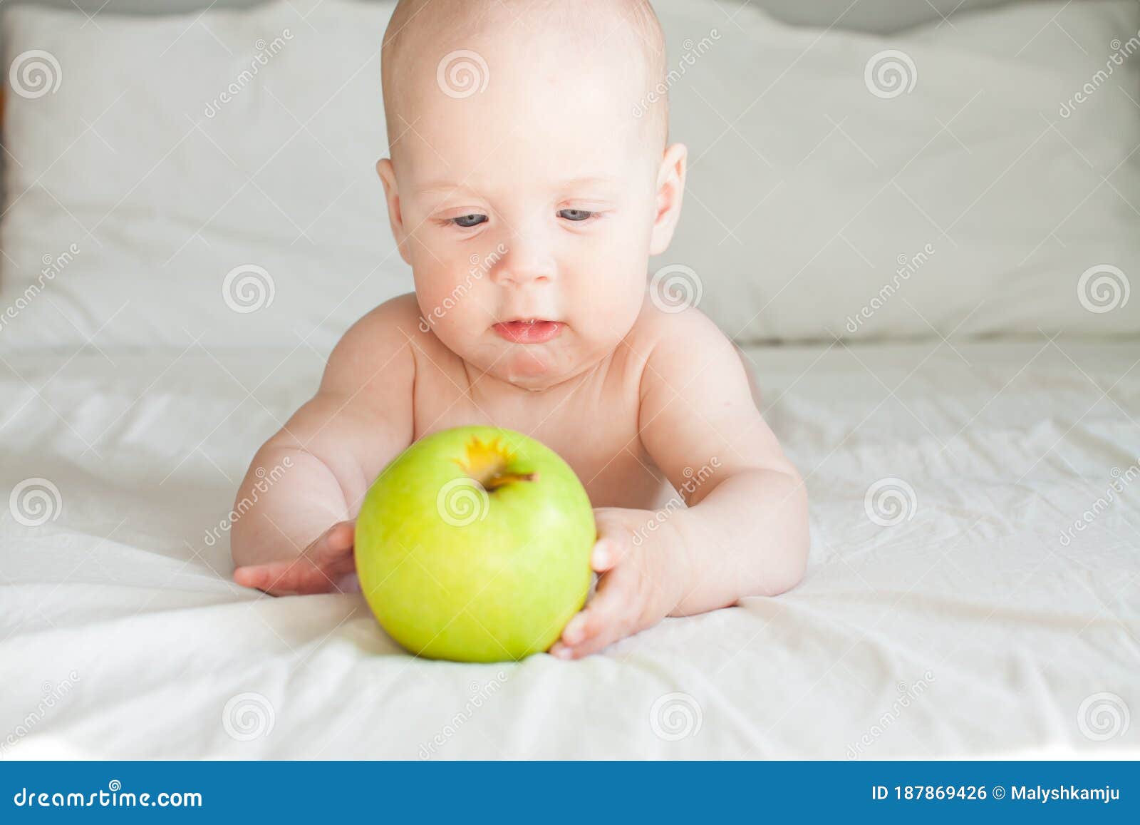 Cute Cheerful Baby Lies on a White Bed and Looks at a Green Apple Lying ... Cute Cheerful Baby Lies on a White Bed and Looks at a Green Apple Lying ...
