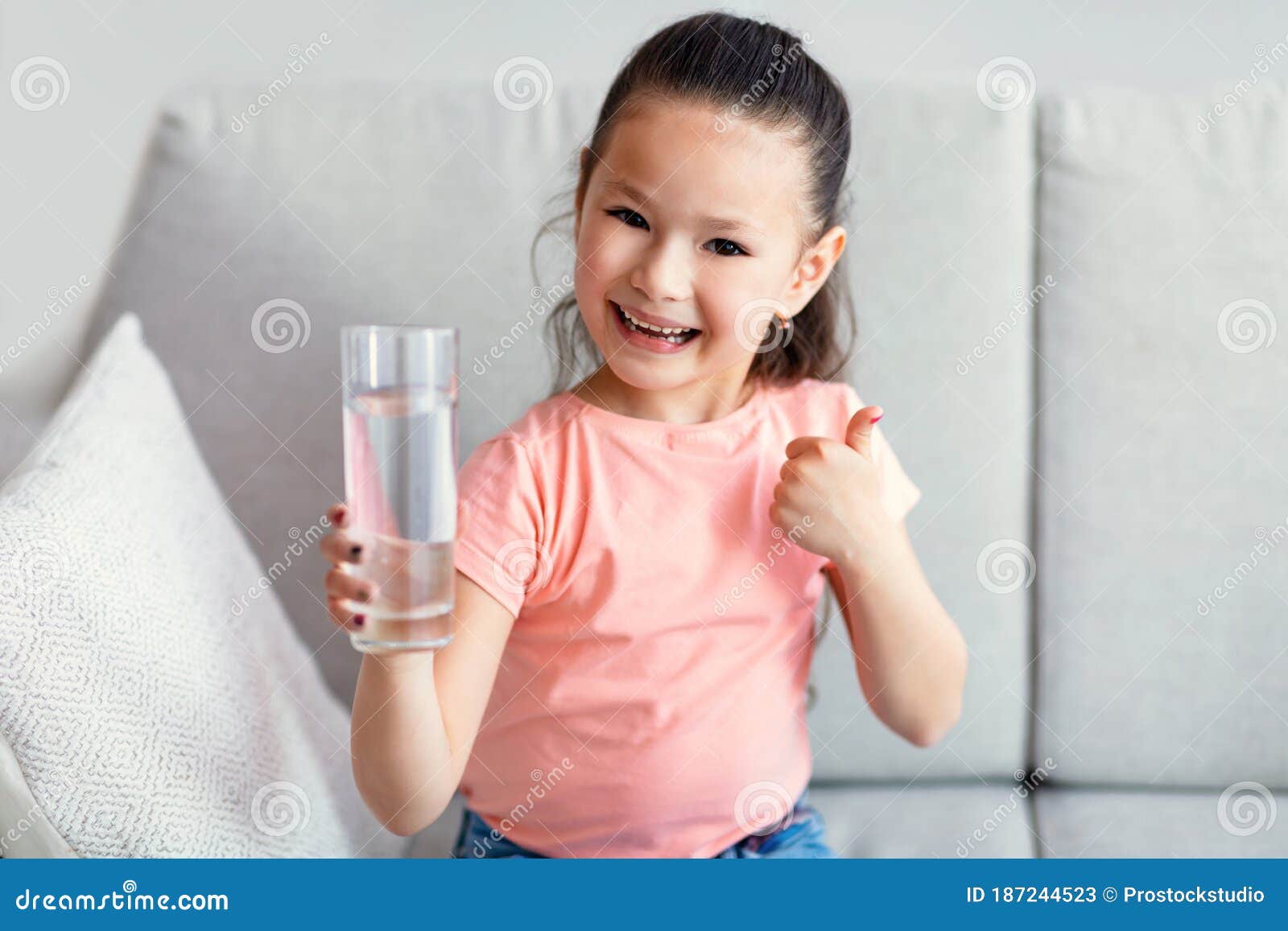 Cute Girl Holding Glass Of Water Gesturing Thumbs Up At Home Stock