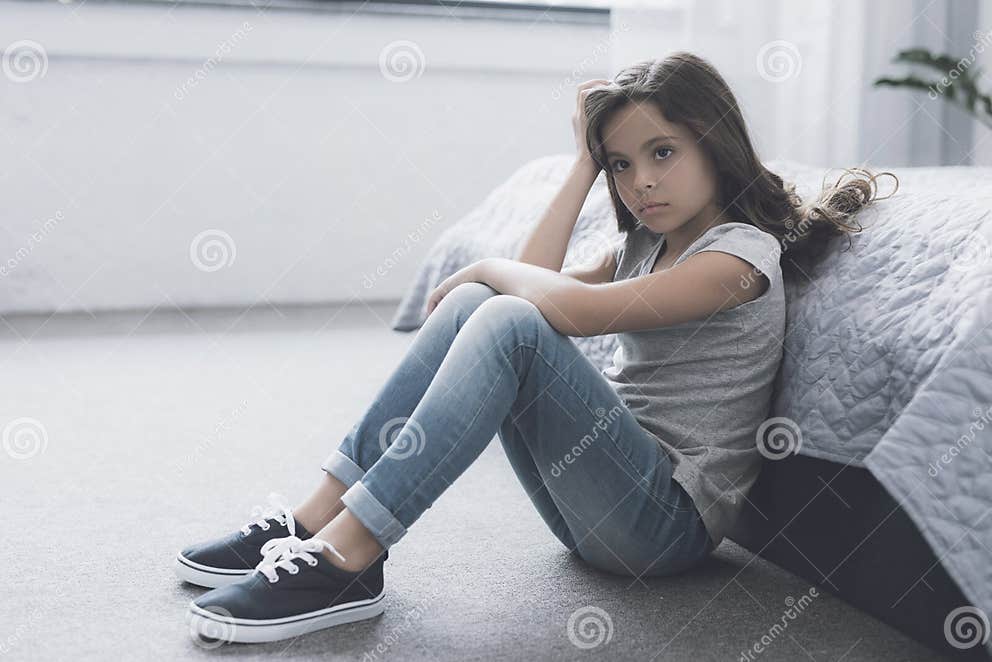 A Small Dark Haired Girl Sits On The Floor Leaning On The Bed And