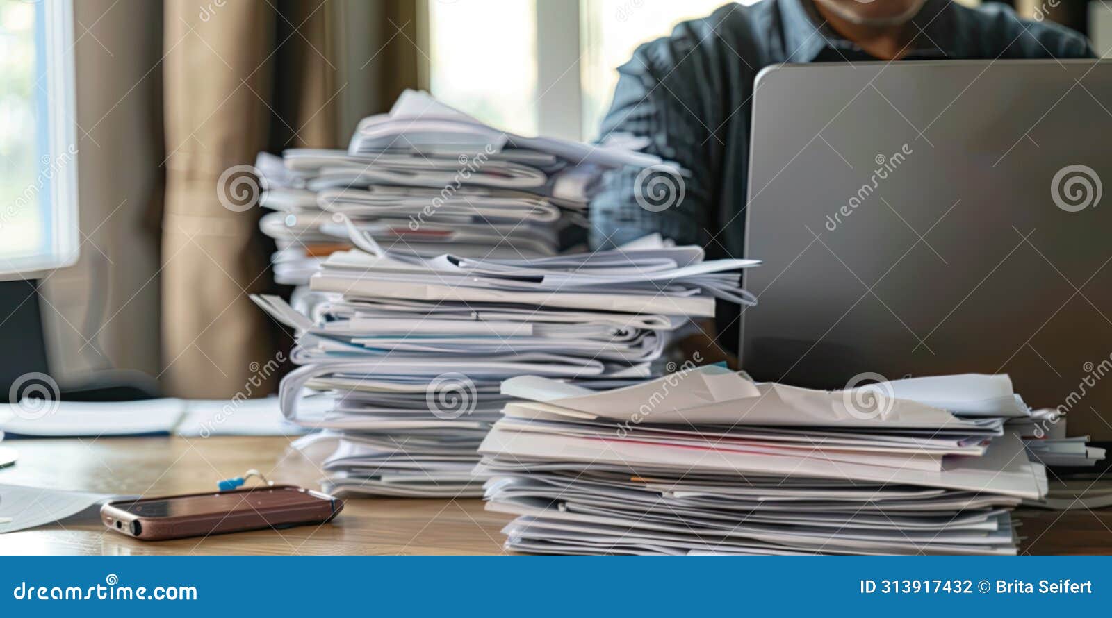 At the Desk Working Person on Laptop Computer with a Pile of Documents ...