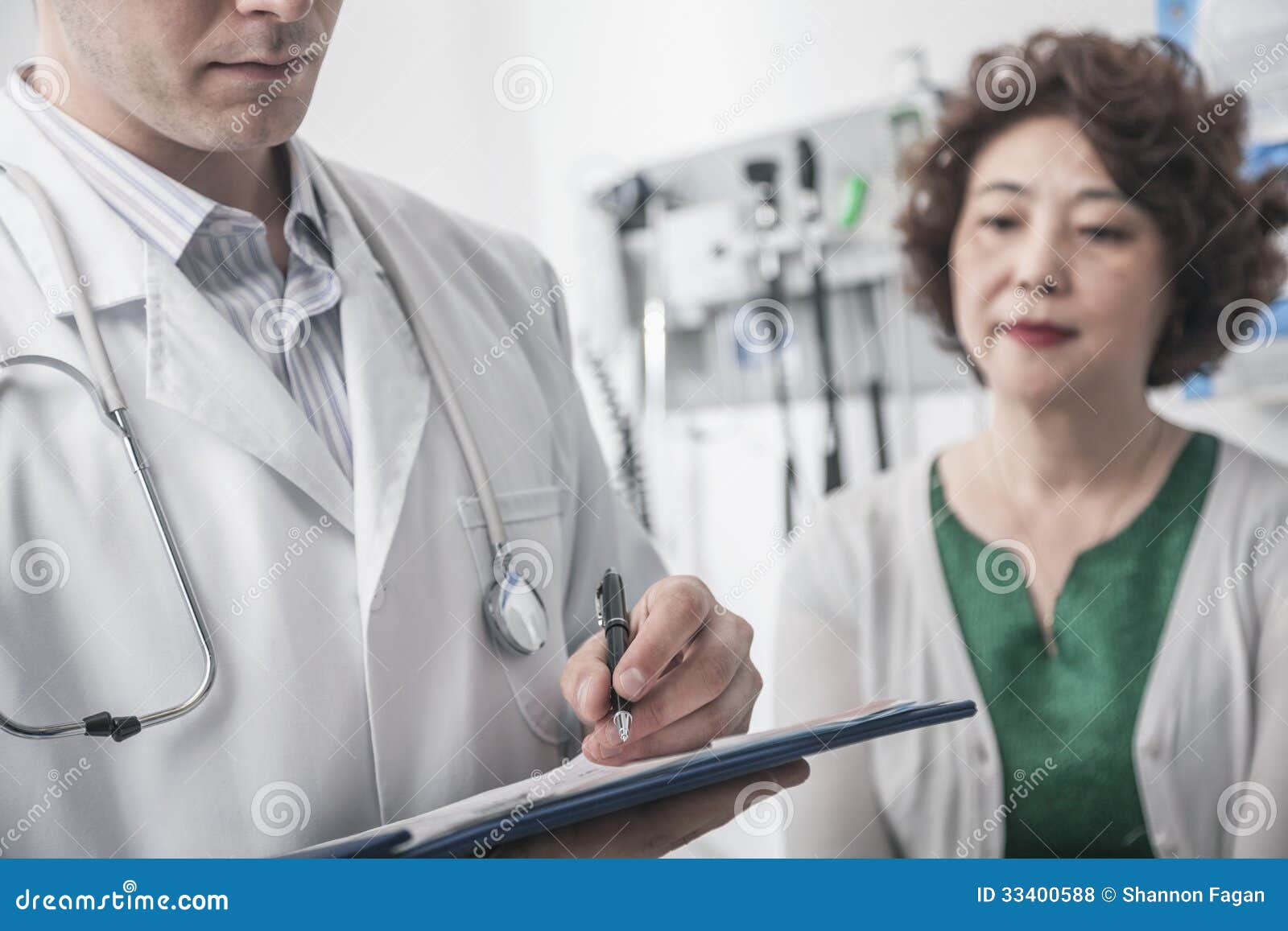 Doctor Writing on Medical Chart with Patient in the Background Stock ...