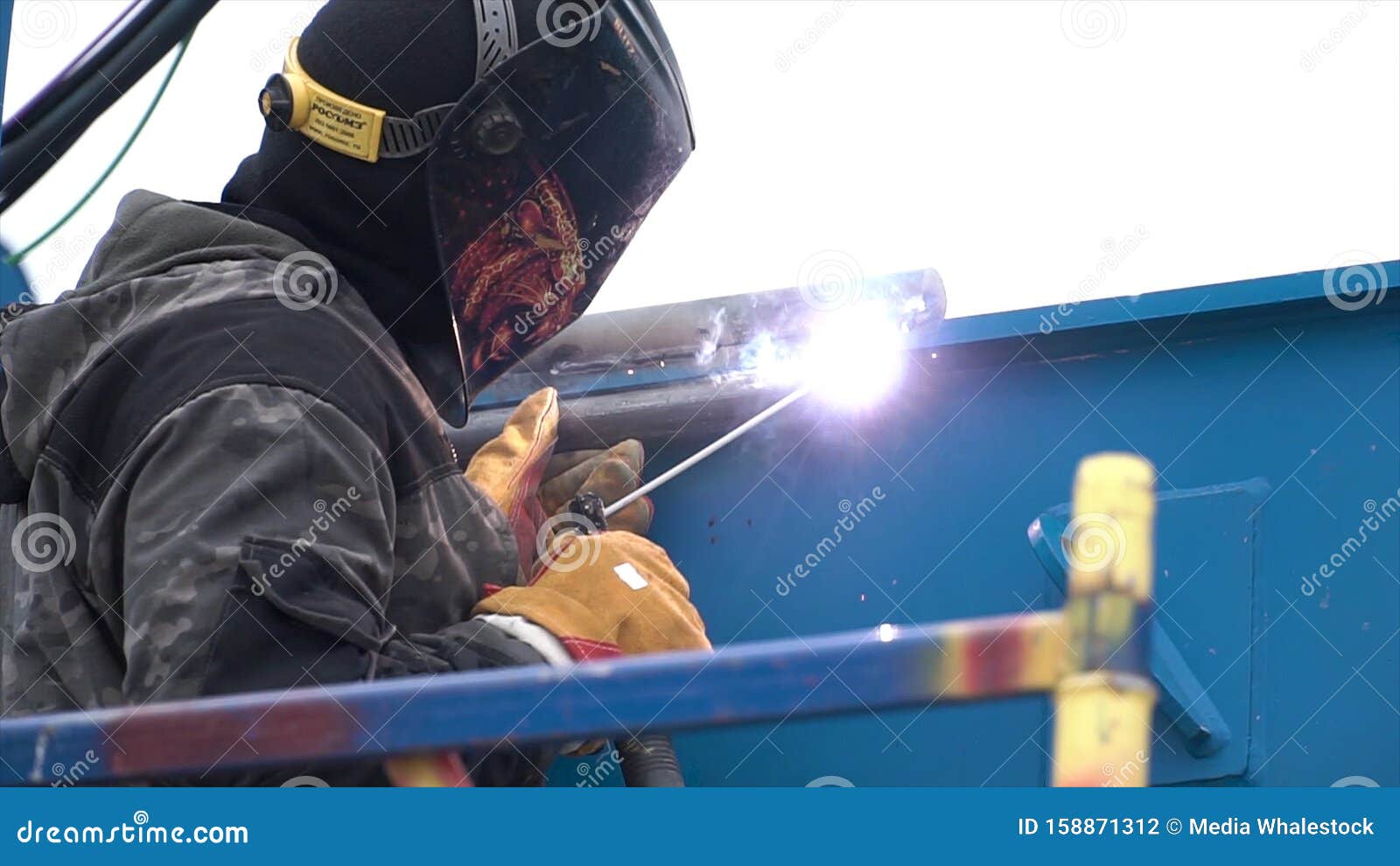Side View of a Man in Protective Welding Mask while Welding Blue Metal ...