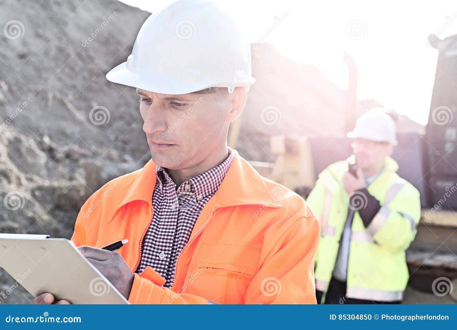 Engineer Writing on Clipboard at Construction Site with Colleague in ...