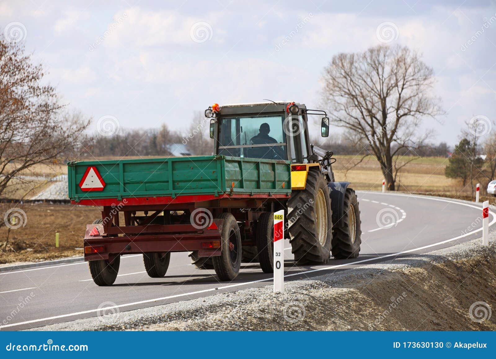 A Farmer on a Tractor Pulls a Trailer Along an Asphalt Road Passing ...