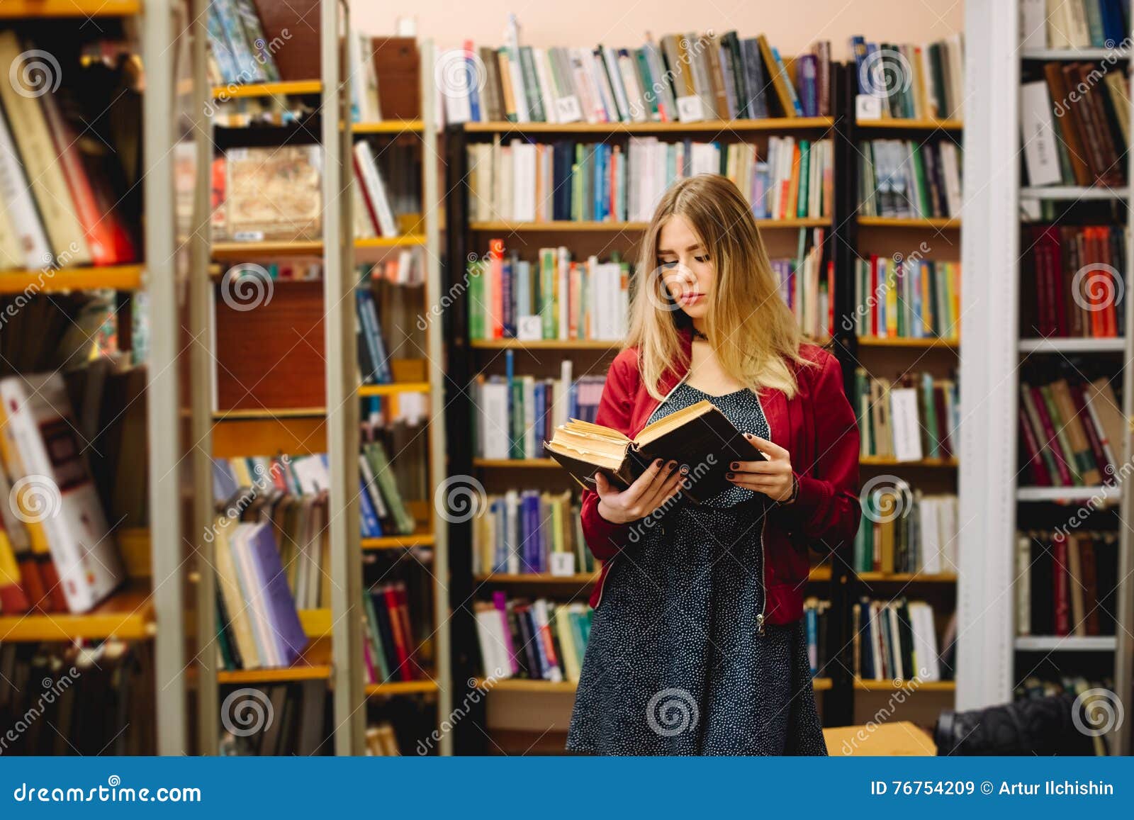 Female Student Reading a Book between Bookshelves in University Library ...