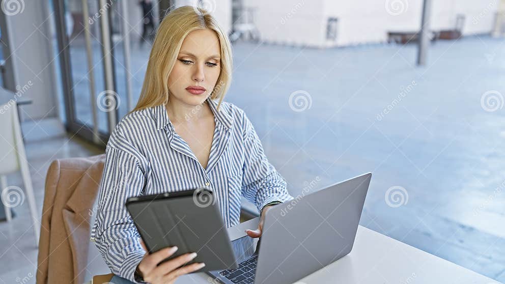 A Focused Blonde Woman Using a Laptop and Tablet Simultaneously in an ...