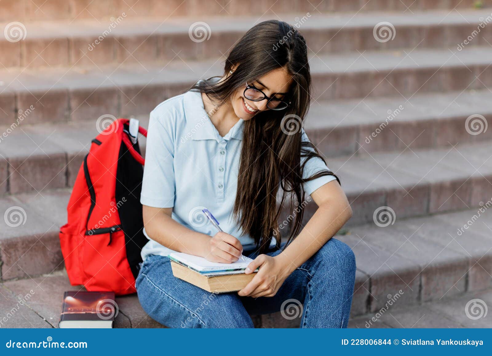 A Focused Student with Glasses, Sitting on the Stairs, Writes in Her ...