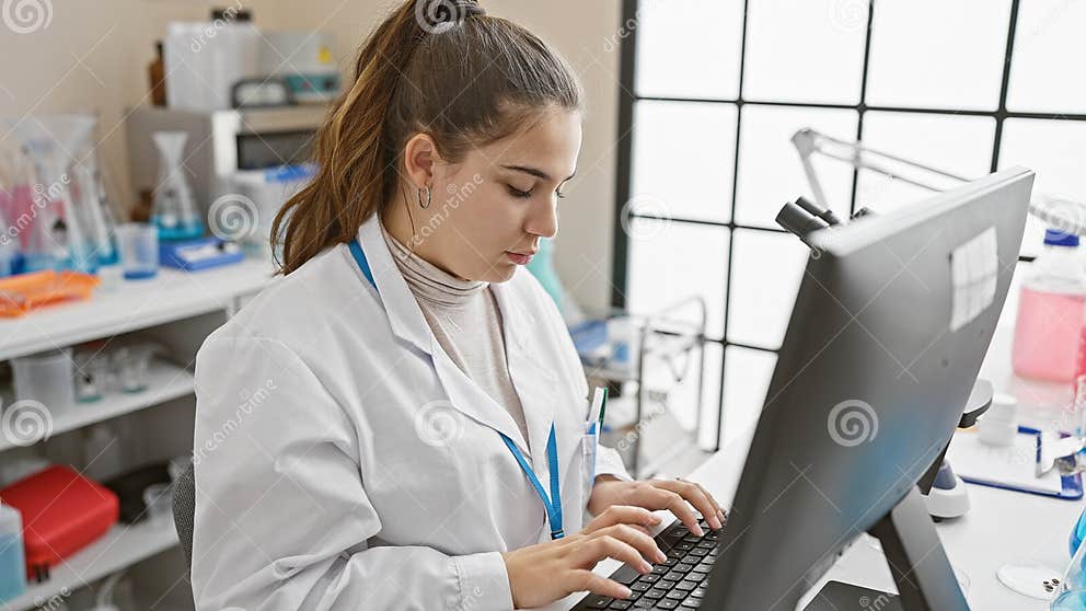 A Focused Young Hispanic Woman in a Lab Coat Typing on a Computer in a ...