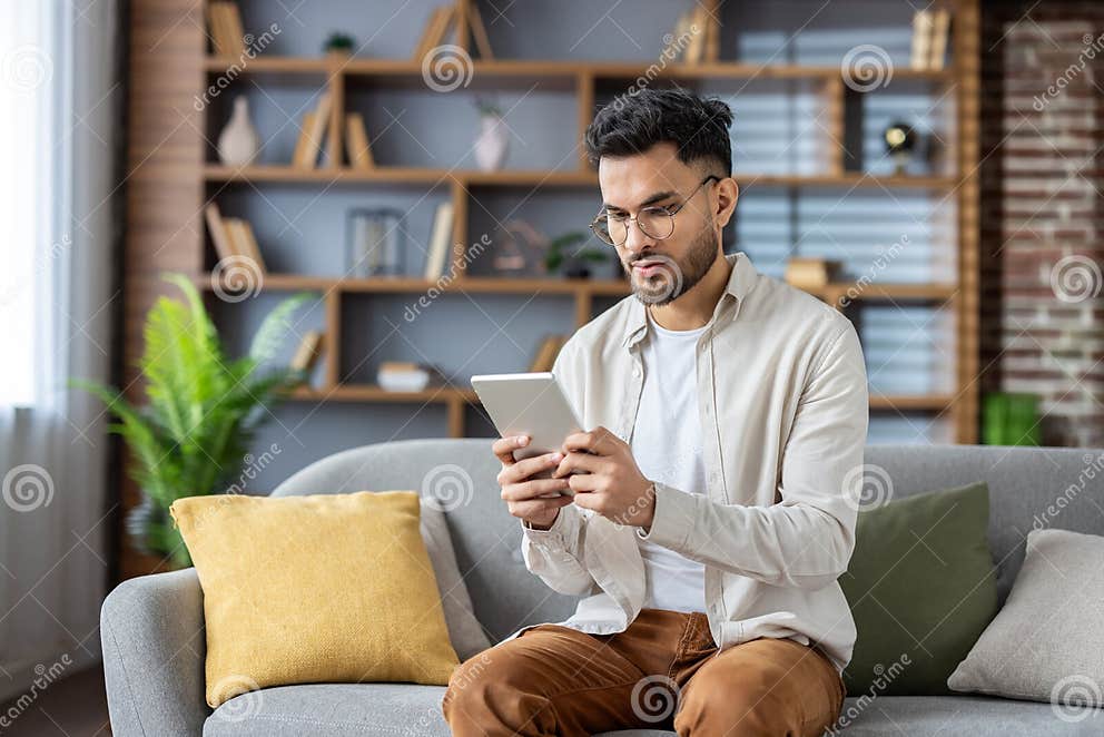 Focused Young Man Reading on Tablet at Home, Comfortably Seated on Sofa ...
