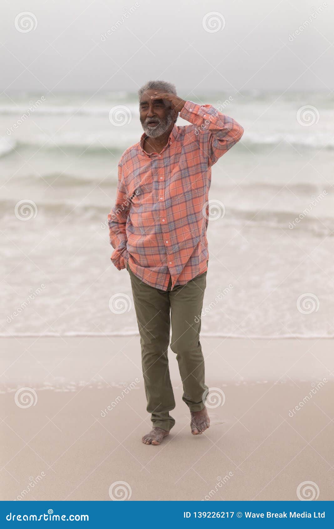 Senior Man with Shielding Eyes and Hand in Pocket Walking on the Beach ...