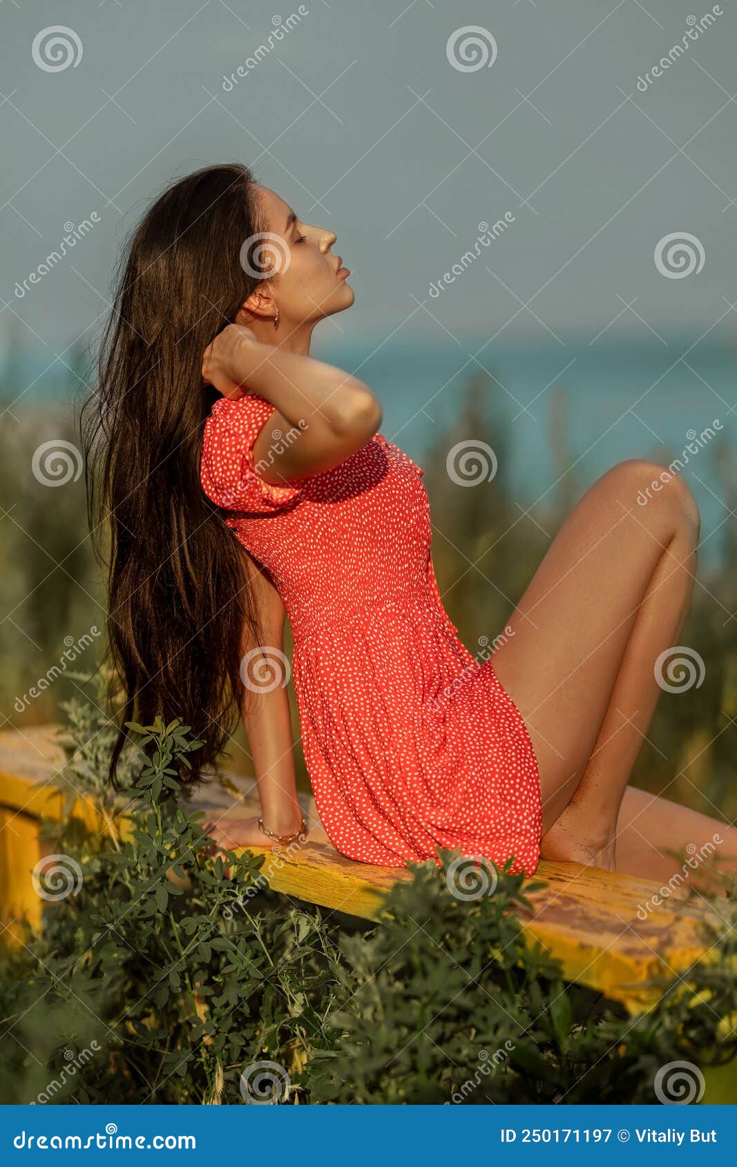 Girl In A Red Dress With Polka Dots Sits On A Bench Overlooking The Sea
