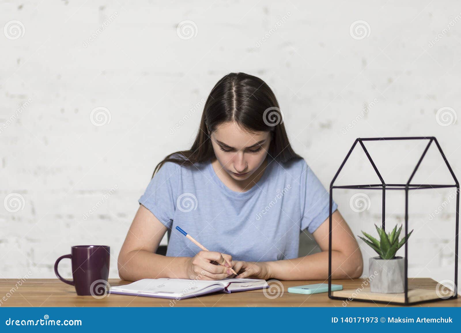The Girl Sits at the Table and Writes in a Notebook with a Pencil Stock ...