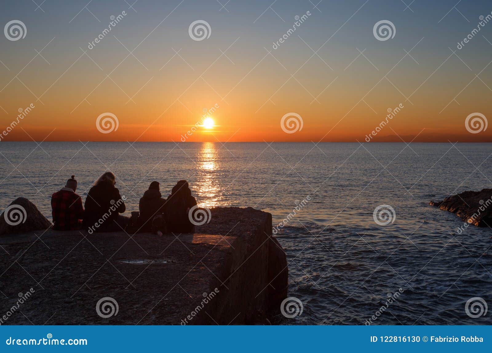 A Group of People Sit Down Looking at Sunset in Front of the Sea ...