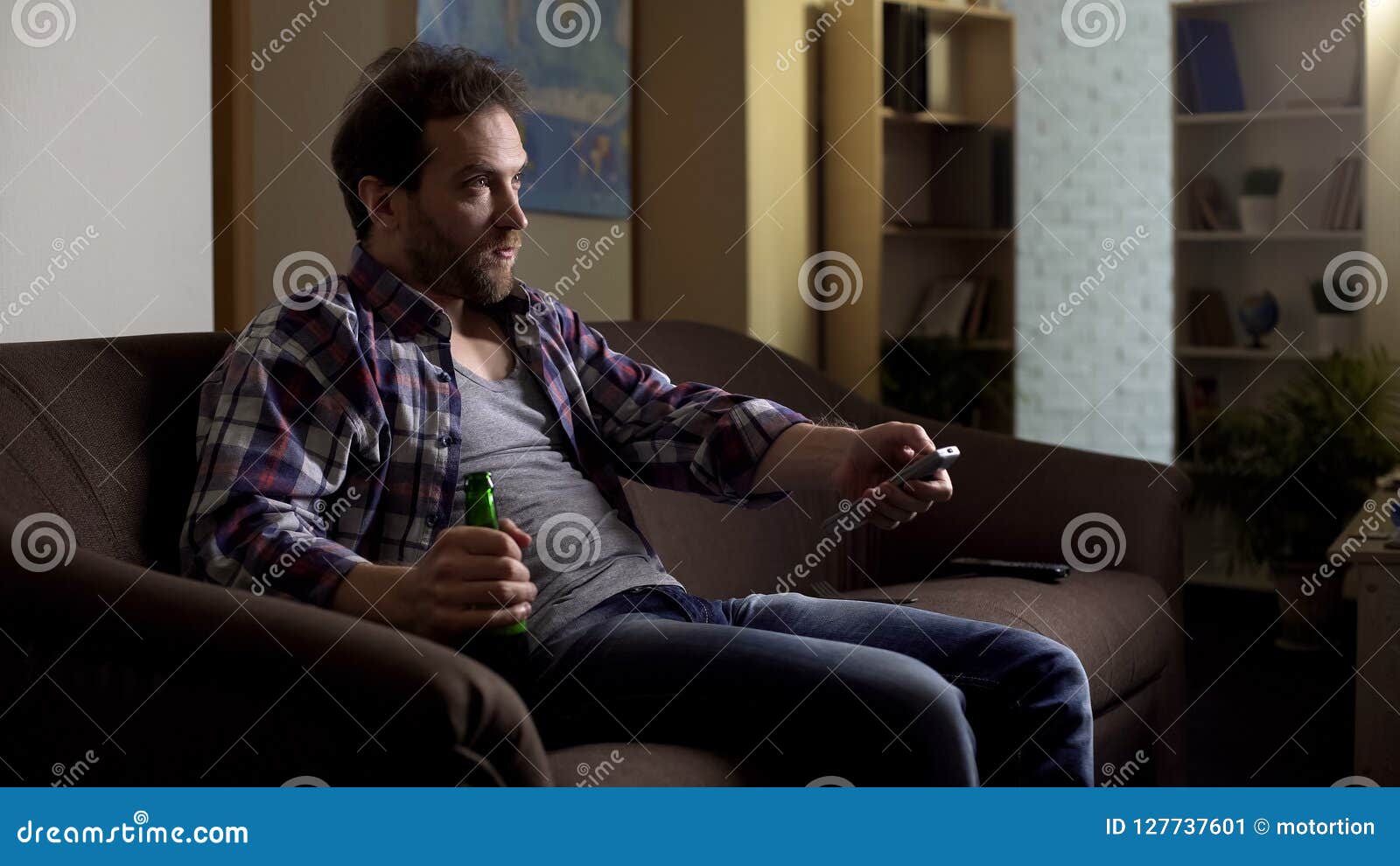 Guy Sitting on Sofa with Beer Bottle in Hand, Using Remote Control To ...