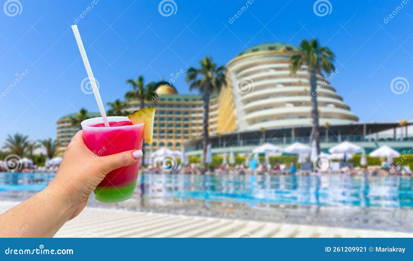 Hand Holding A Glass With Cocktail And Straw And Hotel With Swimming
