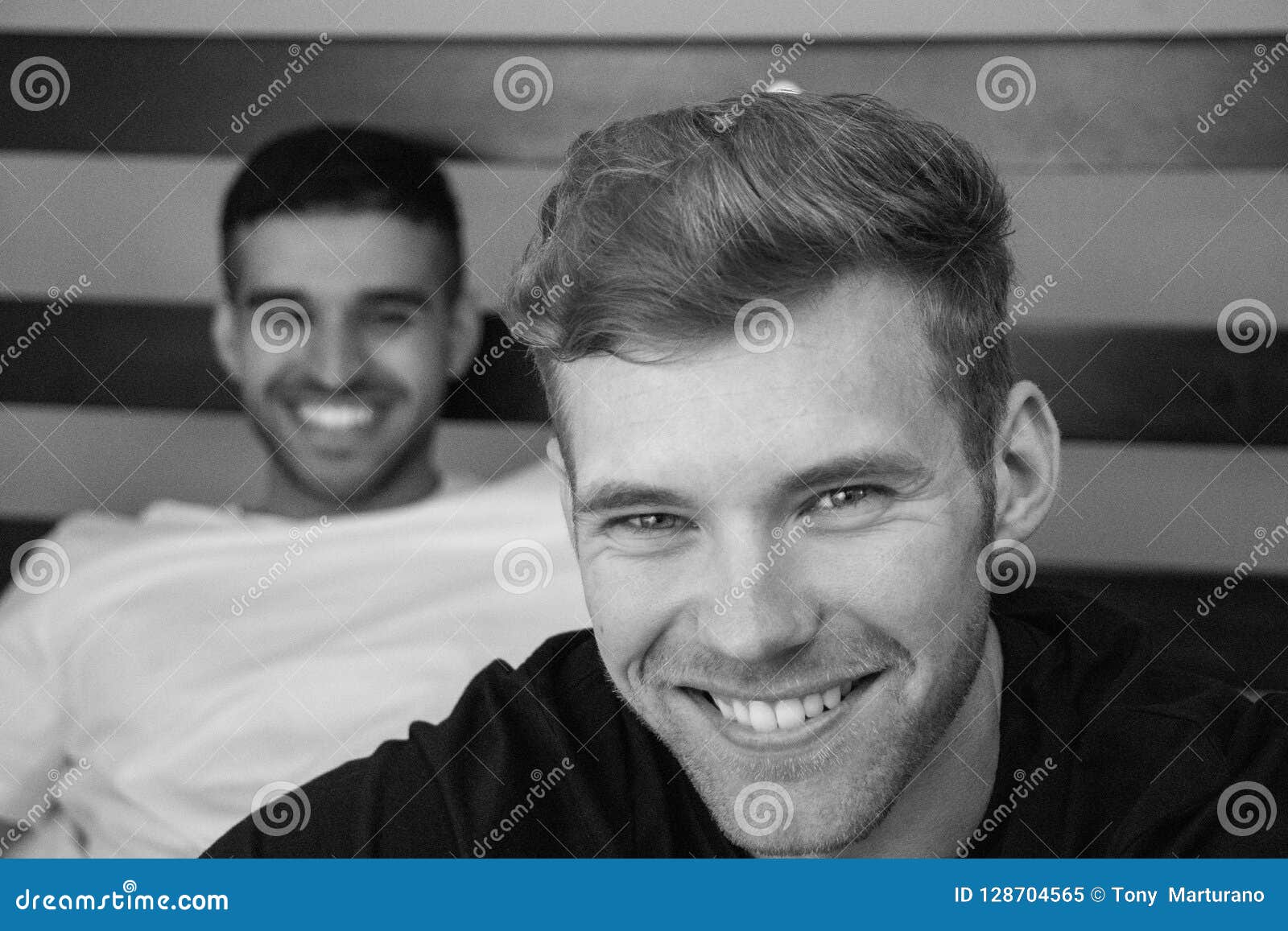 Gay Lovers Sitting In Bed With One Close To Camera And The Other Looking On Both Are Smiling