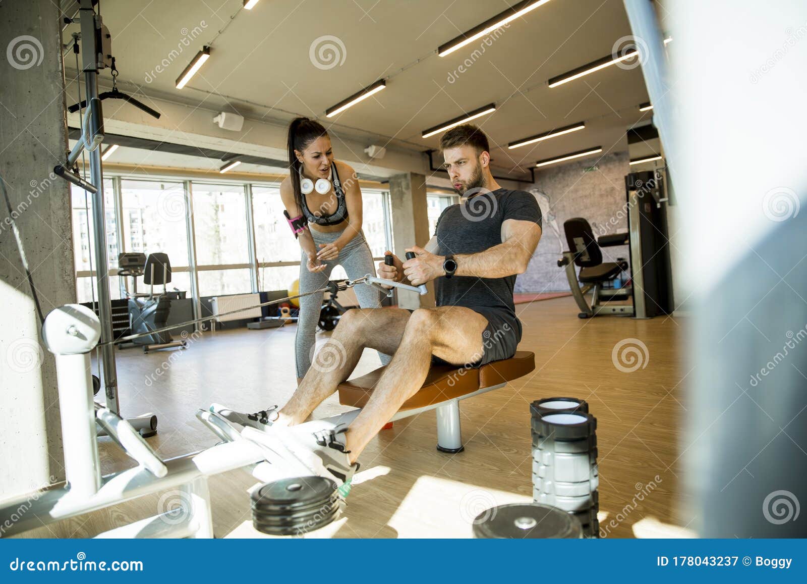 Young Man Using Seated Row Machine in the Gym with Support of Female ...