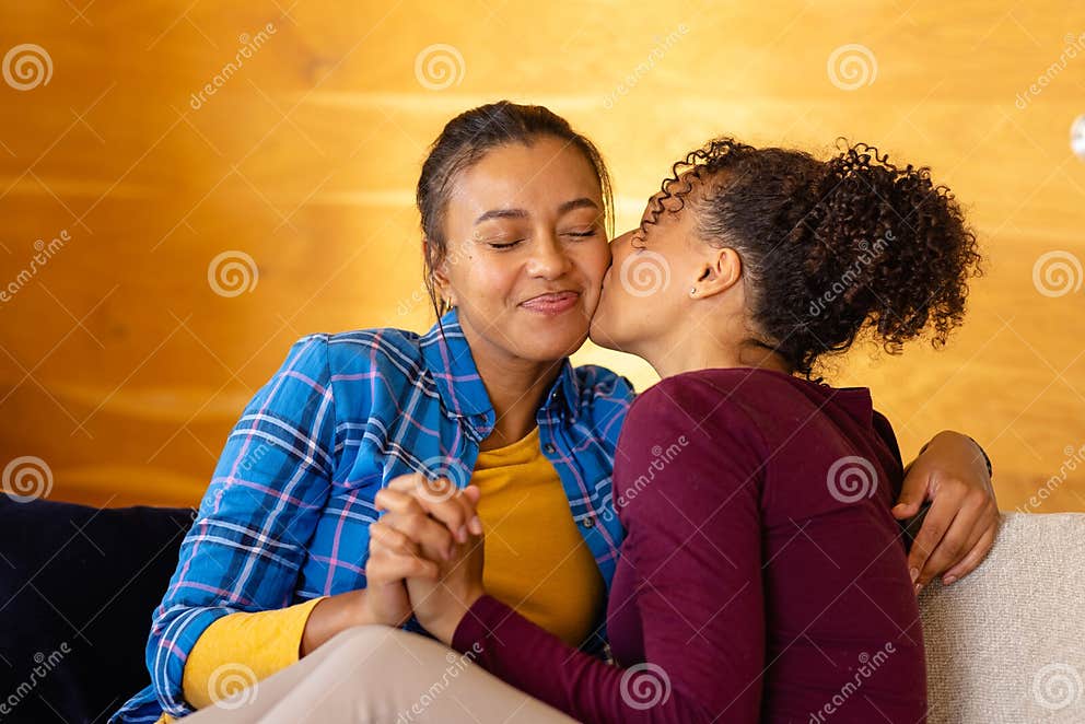 Happy Biracial Lesbian Couple Embracing And Kissing On Sofa In Living