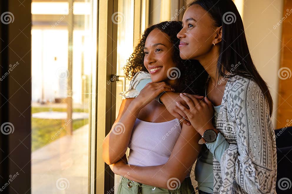 Happy Biracial Lesbian Couple Embracing And Looking Out Window At Home