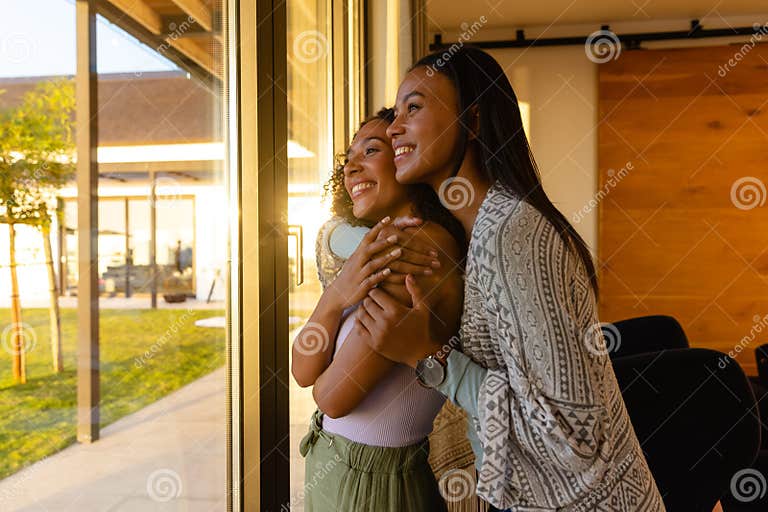 Happy Biracial Lesbian Couple Embracing And Looking Out Window At Home