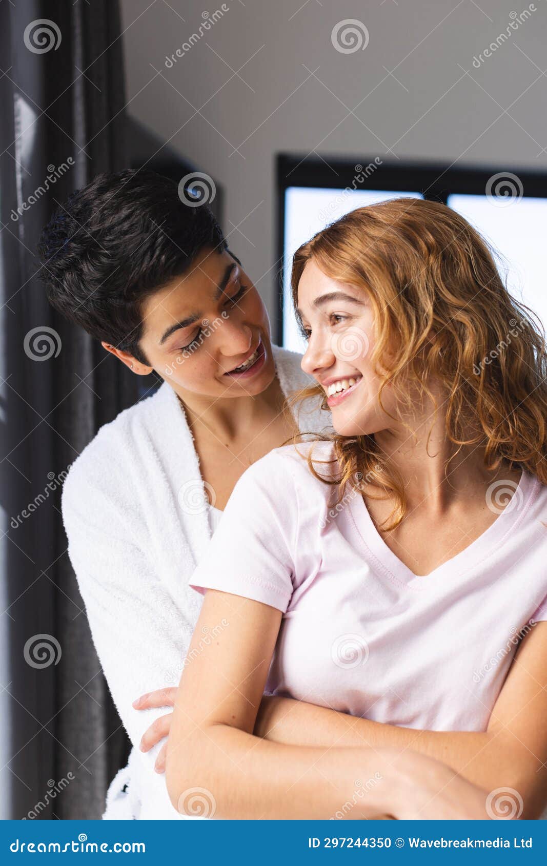 Happy Biracial Lesbian Couple Embracing In Sun By Window At Home Stock