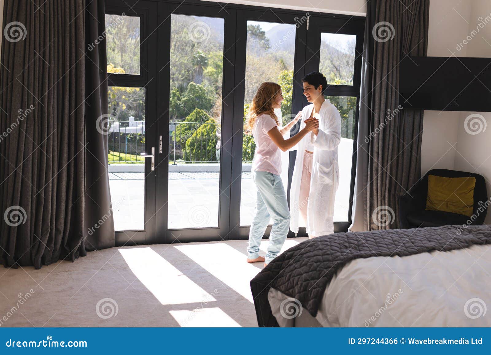 Happy Biracial Lesbian Couple Holding Hands Dancing In Sunny Bedroom At
