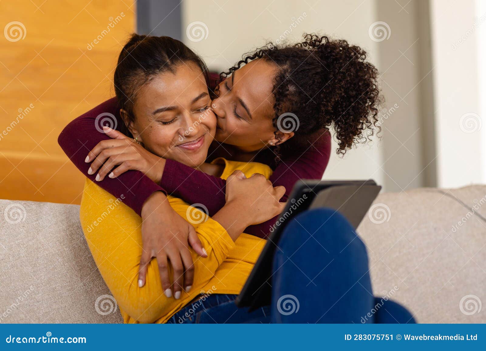 Happy Biracial Lesbian Couple Hugging And Using Tablet On Sofa In