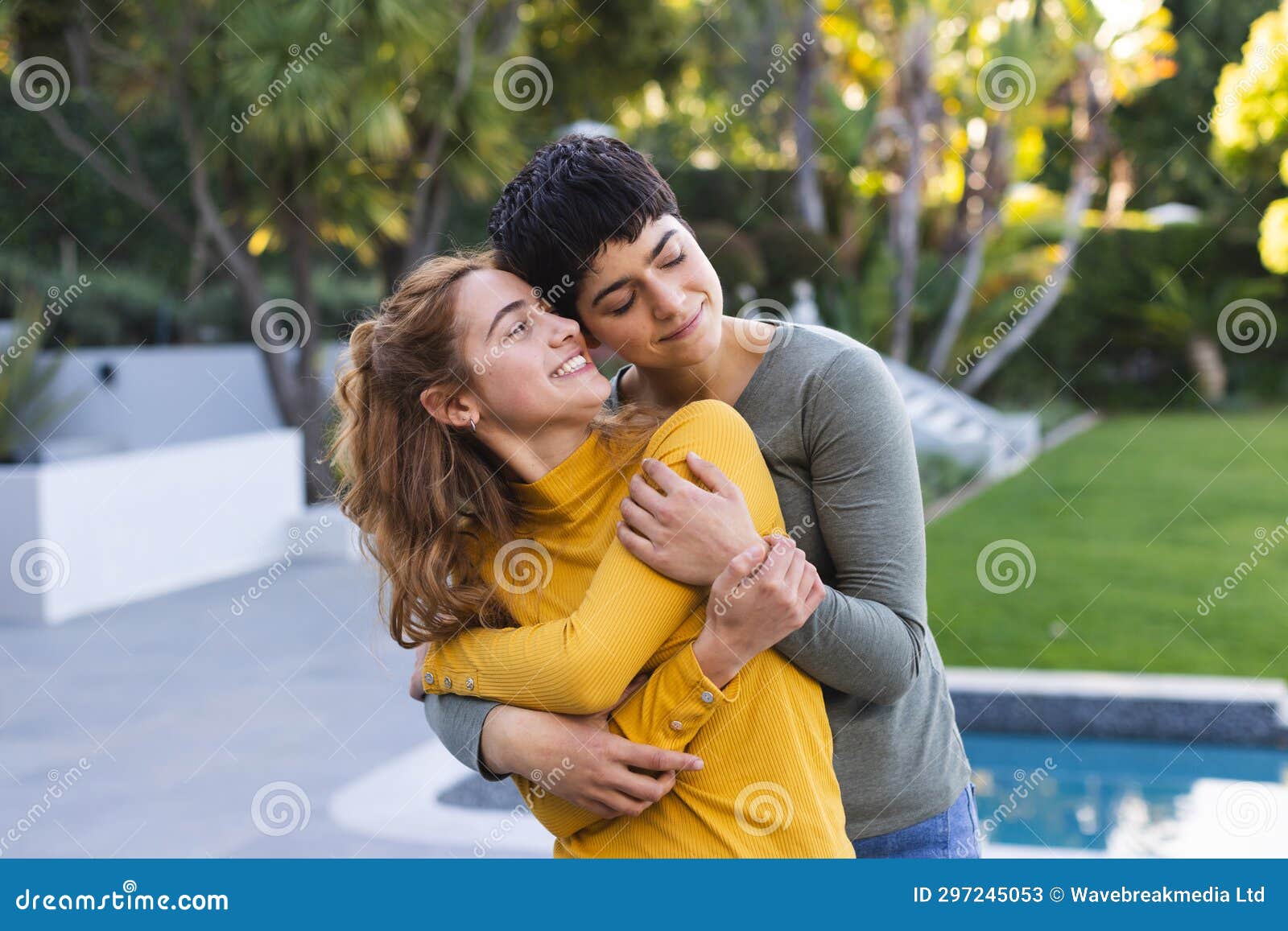 Happy Biracial Lesbian Couple Standing On Garden Terrace By Pool