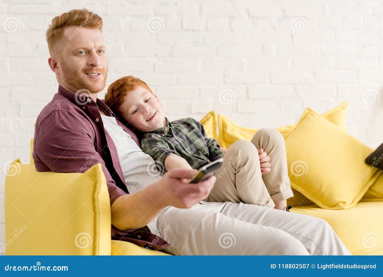 Happy Father And Son Sitting Together On Sofa And Using Remote