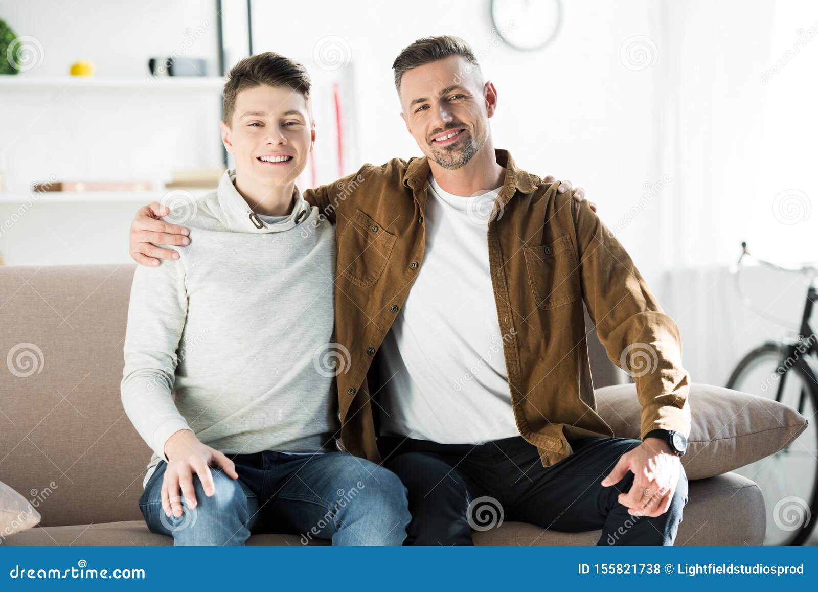 Happy Father And Teen Son Sitting On Sofa And Looking At Camera Stock