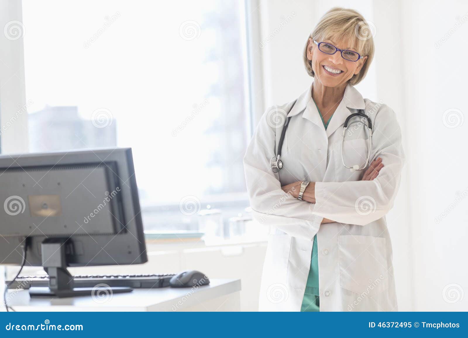 Happy Mature Female Doctor Standing Arms Crossed In Hospital Stock