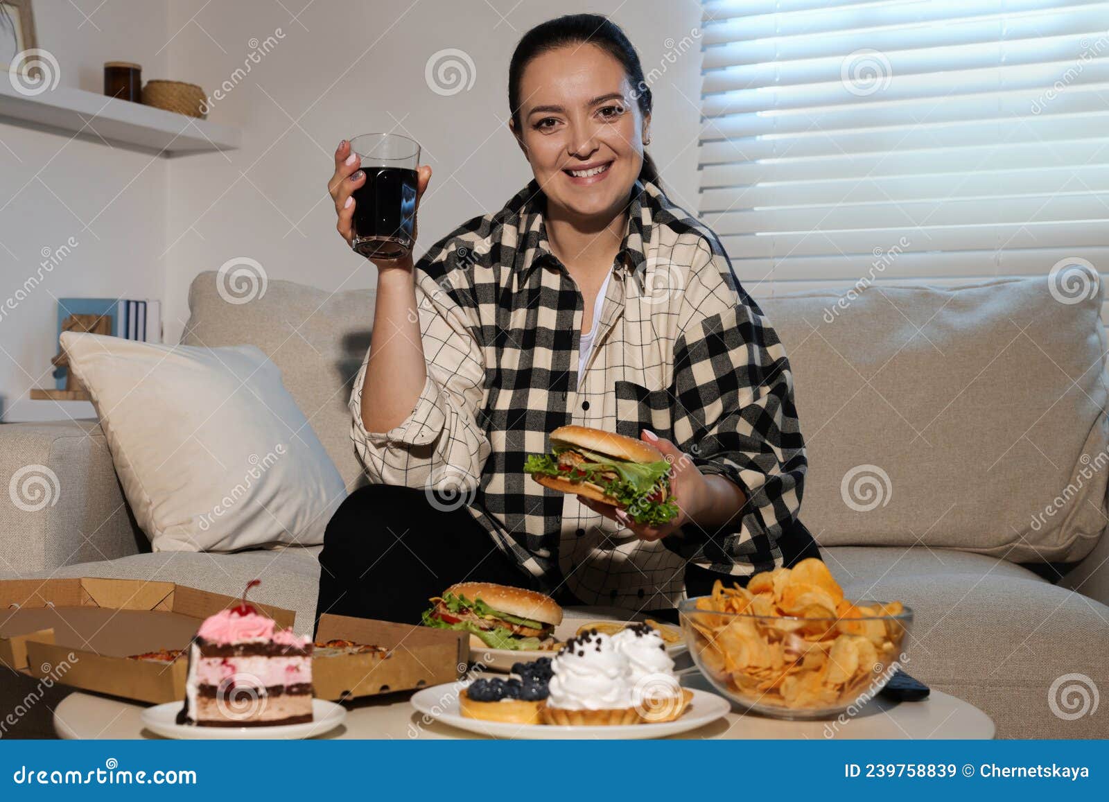 Happy Overweight Woman With Glass Of Cola And Burger On Sofa At Home