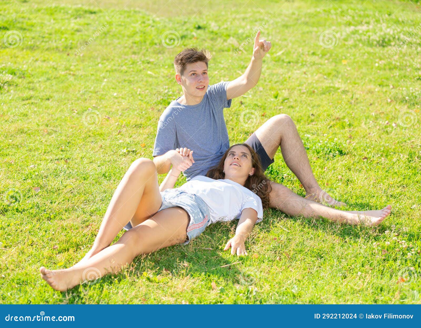 Happy Romantic Couple Sitting On Grass And Points To Something In Day