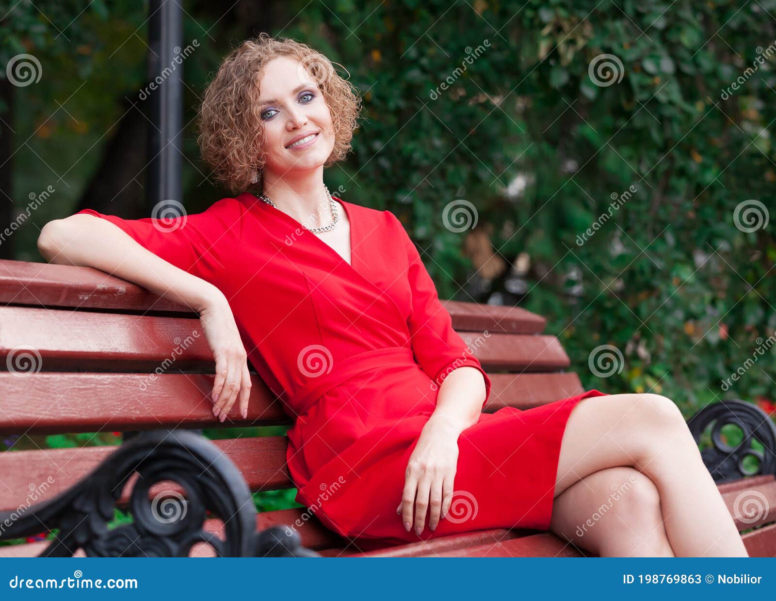Happy Woman In Red Dress Is Sitting On A Bench In A Park And Smiling To You Stock Image Image