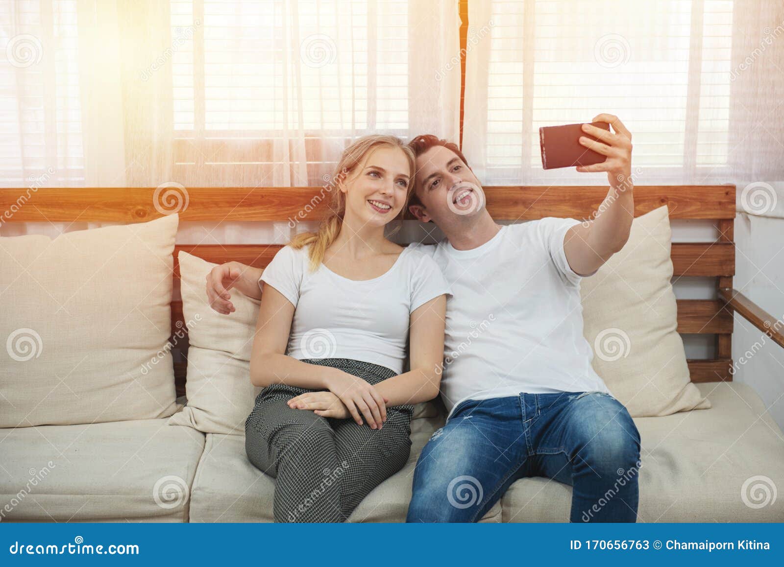 Happy Young Caucasian Couple Sit On The Couch Taking Selfie At Home In