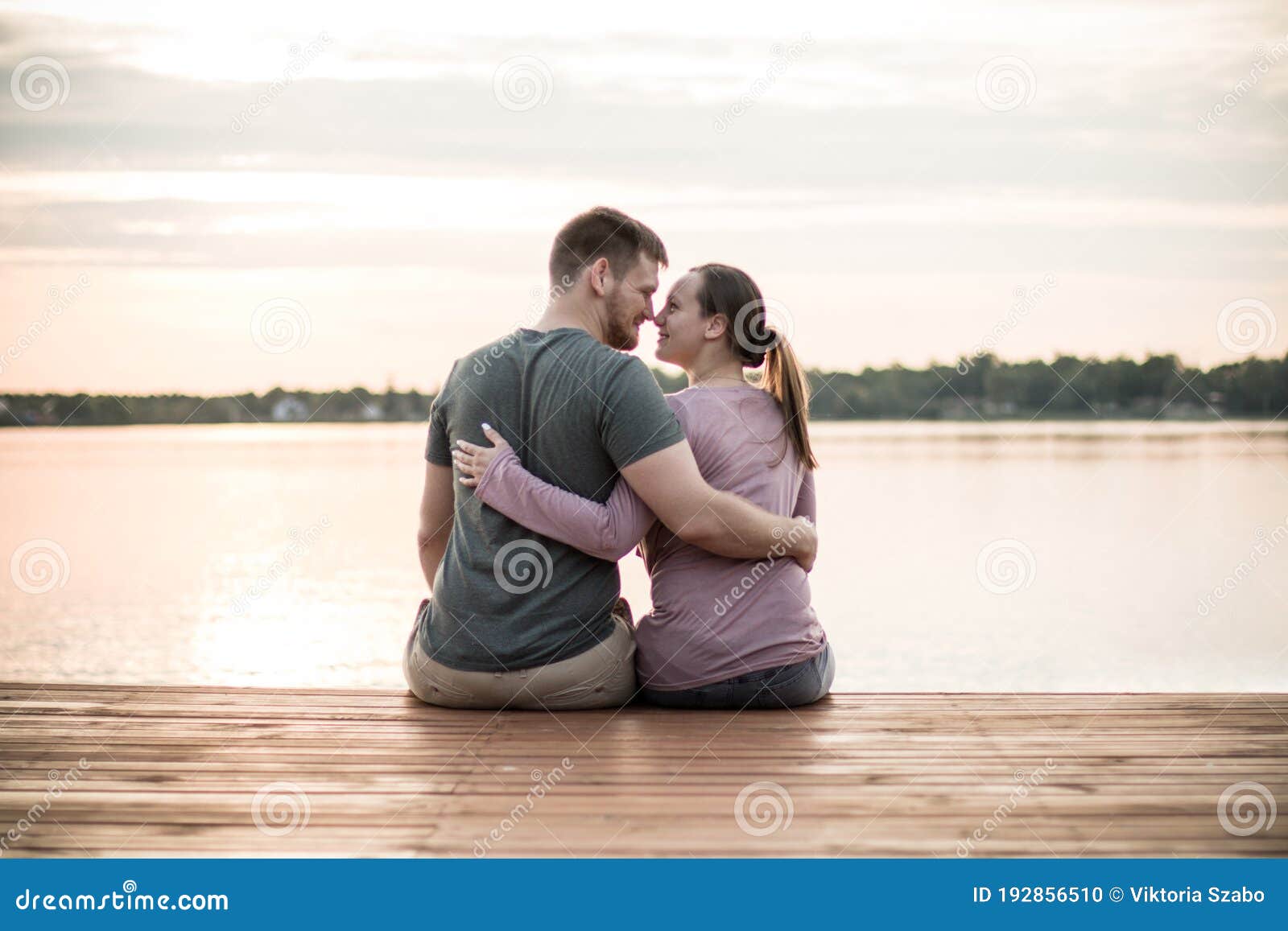 Happy Young Couple Sitting On The Dock By The Lake Hugging Royalty