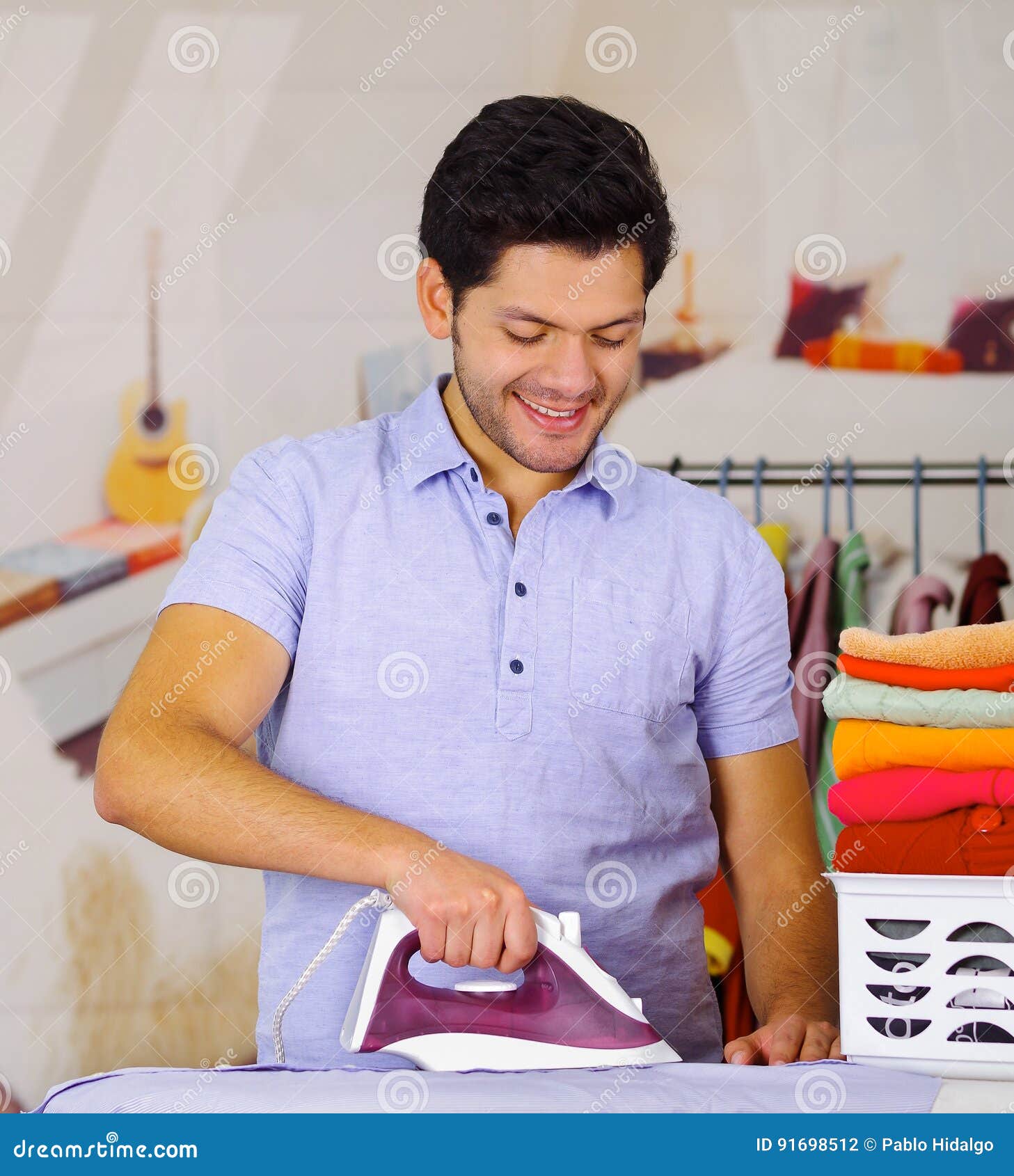 Happy Young Man Ironing Clothes on Ironing Board while he is Using His ...