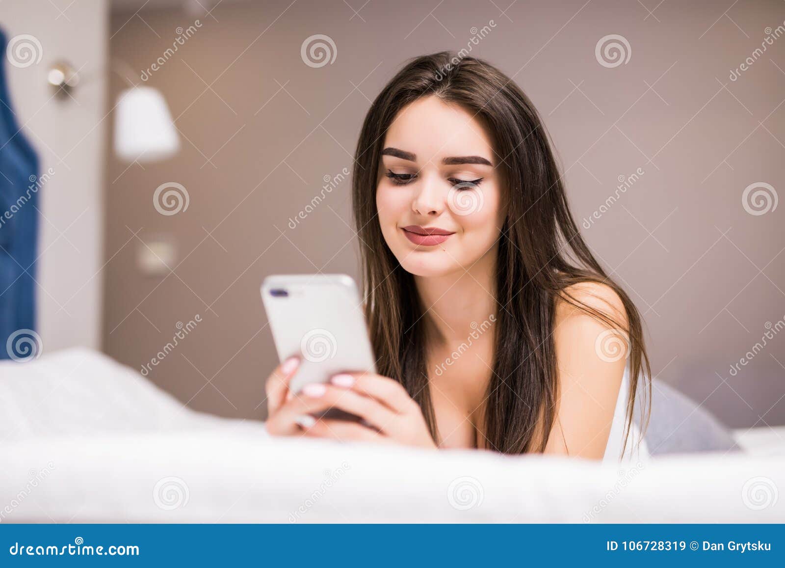 Happy Young Woman Lying In Bed And Texting On Smartphone At Home