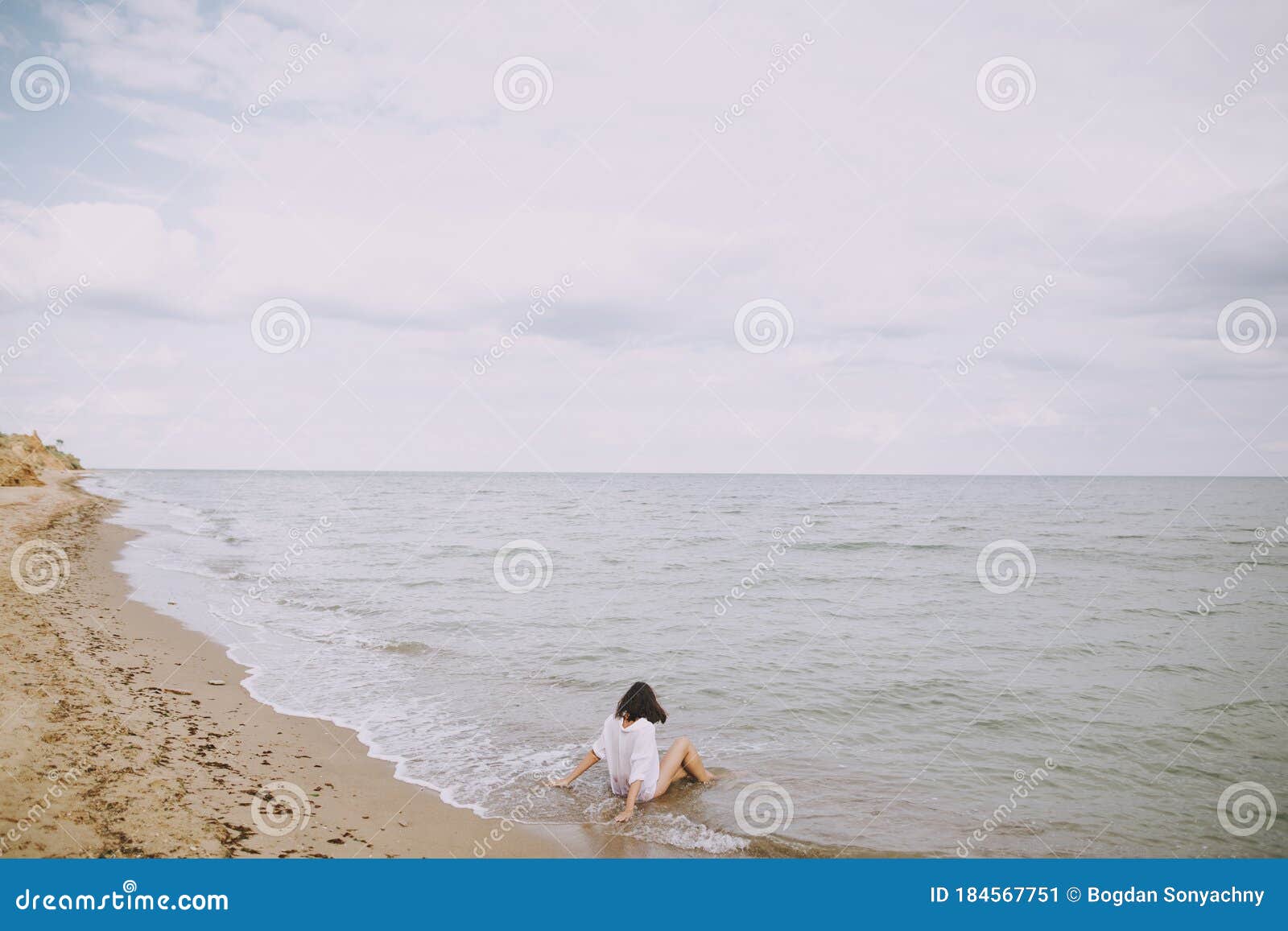 Happy Young Woman In White Shirt Sitting On Beach In Splashing Waves