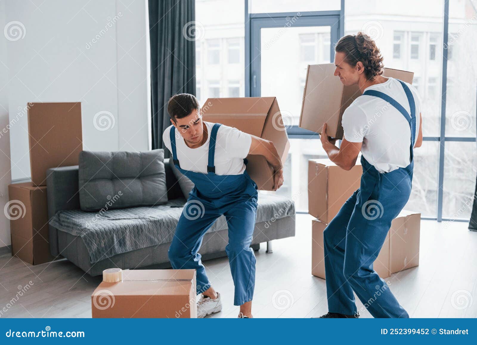 Heavy Boxes. Two Young Movers in Blue Uniform Working Indoors in the ...