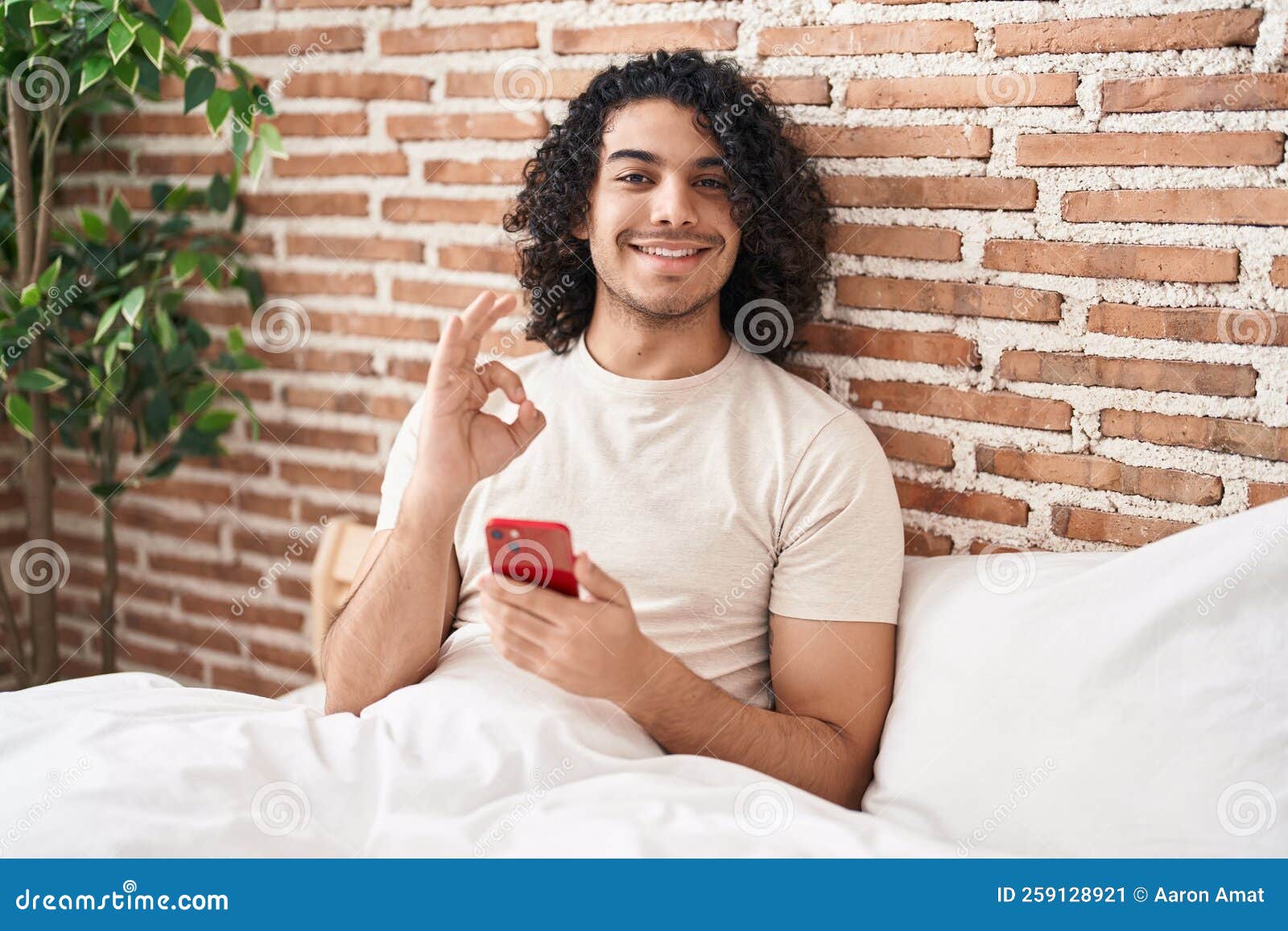 Hispanic Man With Curly Hair Using Smartphone Sitting On The Bed Doing