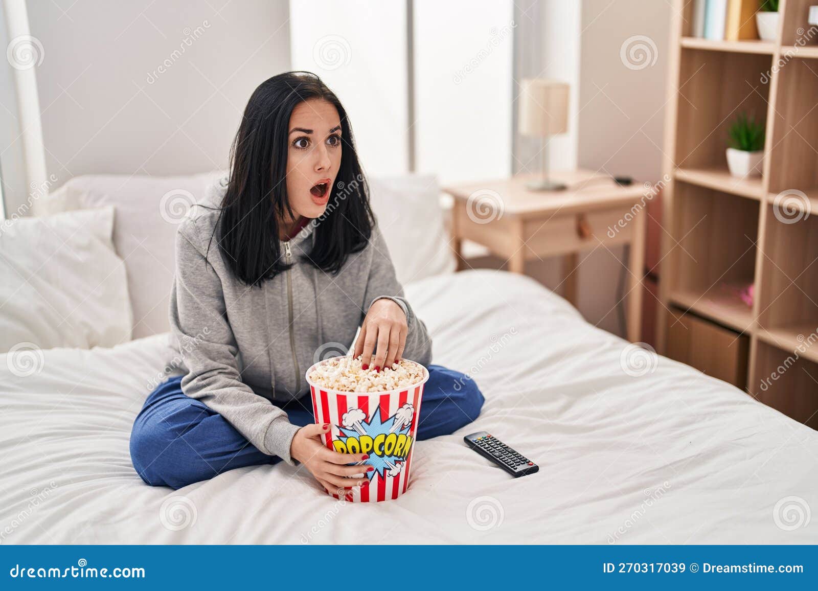 Hispanic Woman Eating Popcorn Watching A Movie On The Bed In Shock Face