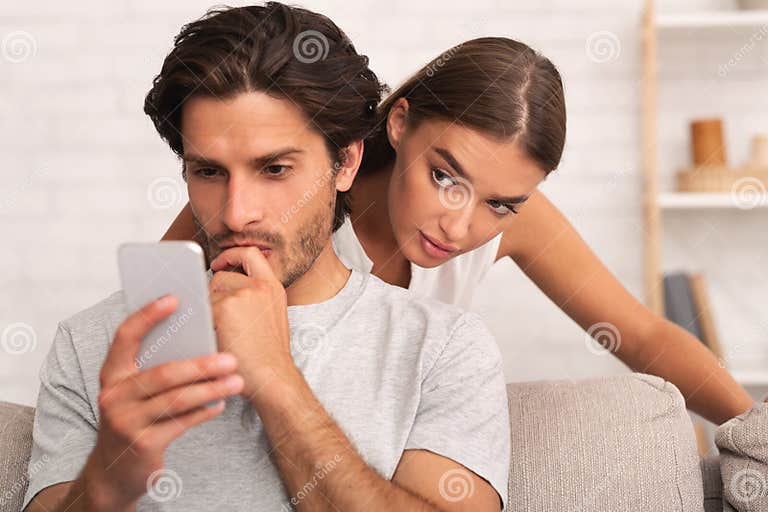 Jealous Wife Spying On Husband While He Texting On Phone Indoor Stock