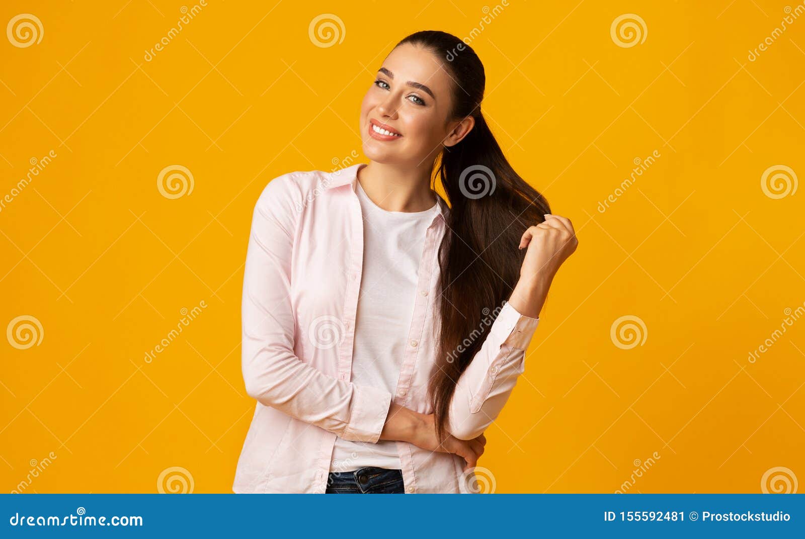 Smiling Young Woman Touching Hair Posing On Yellow Background Stock