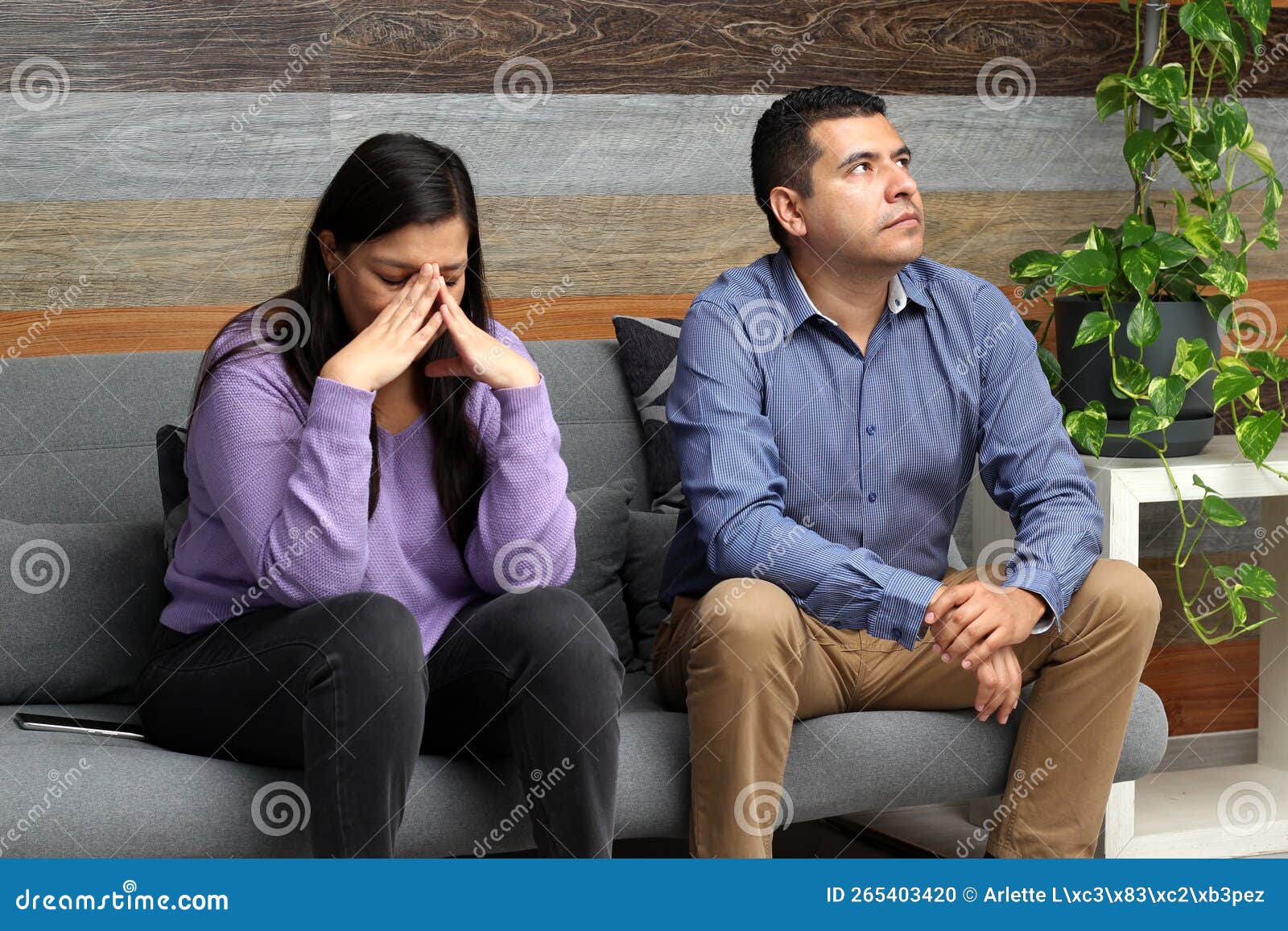 Latino Adult Couple Of Man And Woman Sitting On A Sofa Are Upset And
